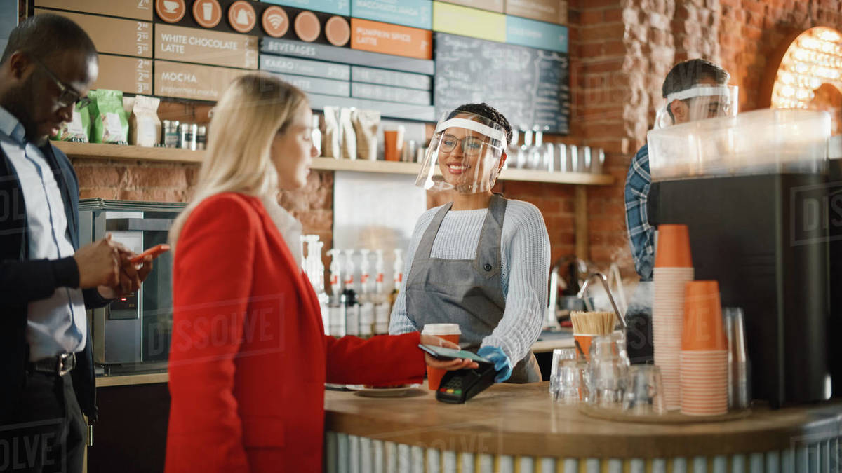Close-up view of barista in a face shield handing payment terminal to ...