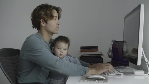 Man sitting at home office desk typing on computer while watching baby ...