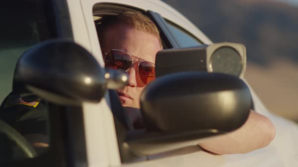 Policeman sitting in police car leaning out window pointing radar gun ...