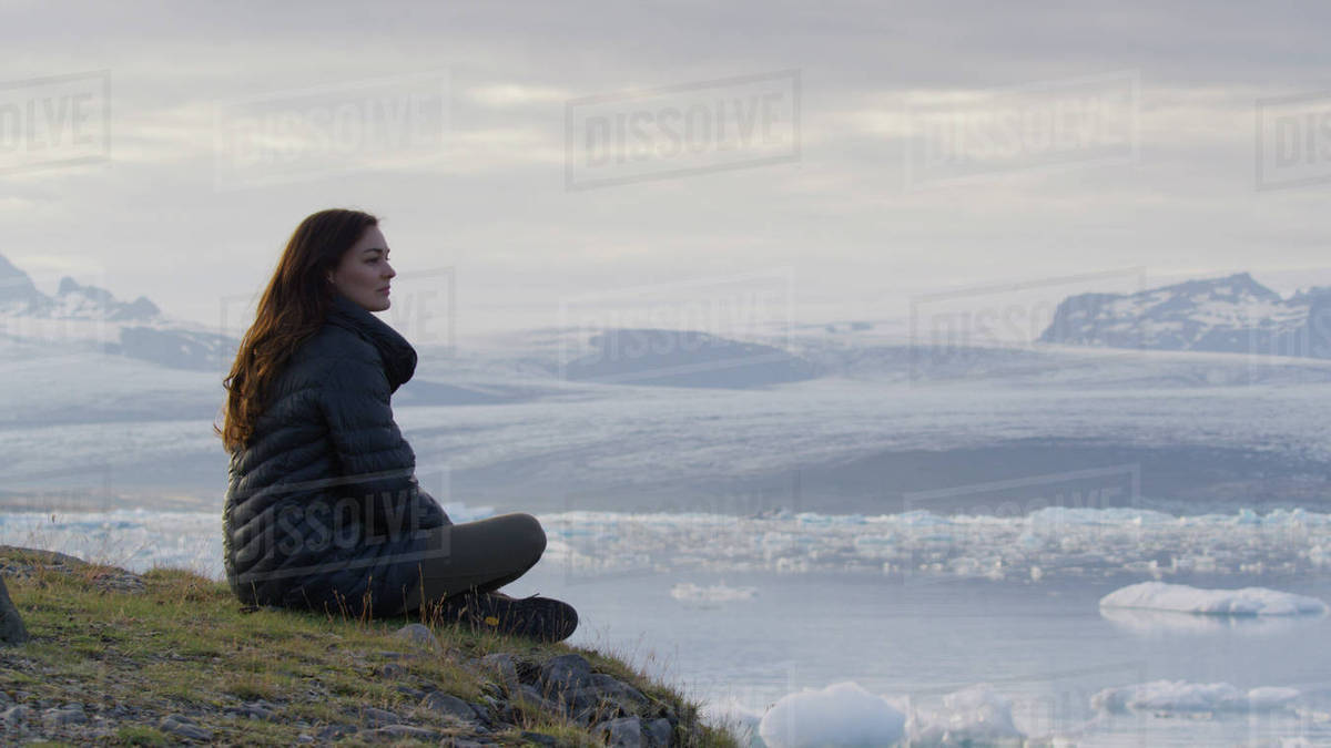 Profile view of woman sitting on grassy hilltop overlooking landscape ...
