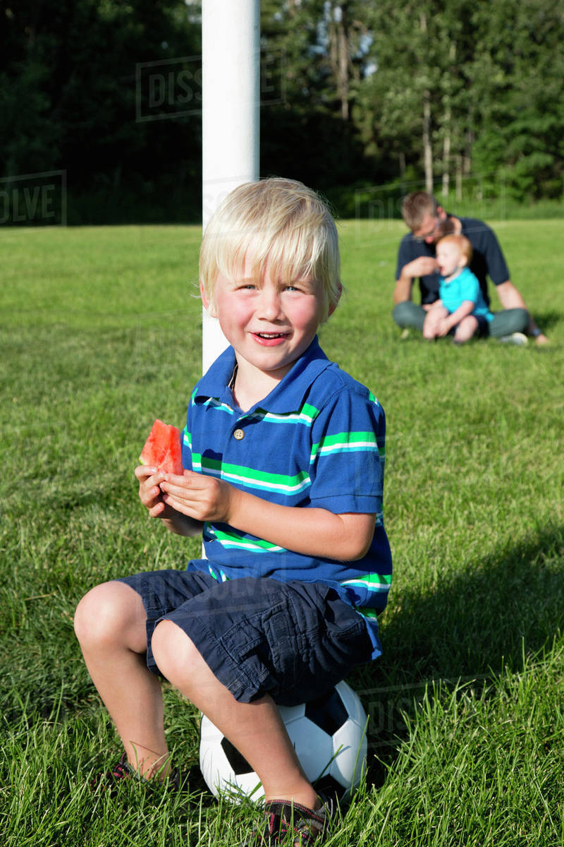 A young boys sits on a soccer ball eating a piece of watermelon in a ...