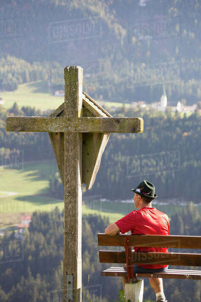 Male hiker sitting on bench with large wooden cross overlooking valley ...