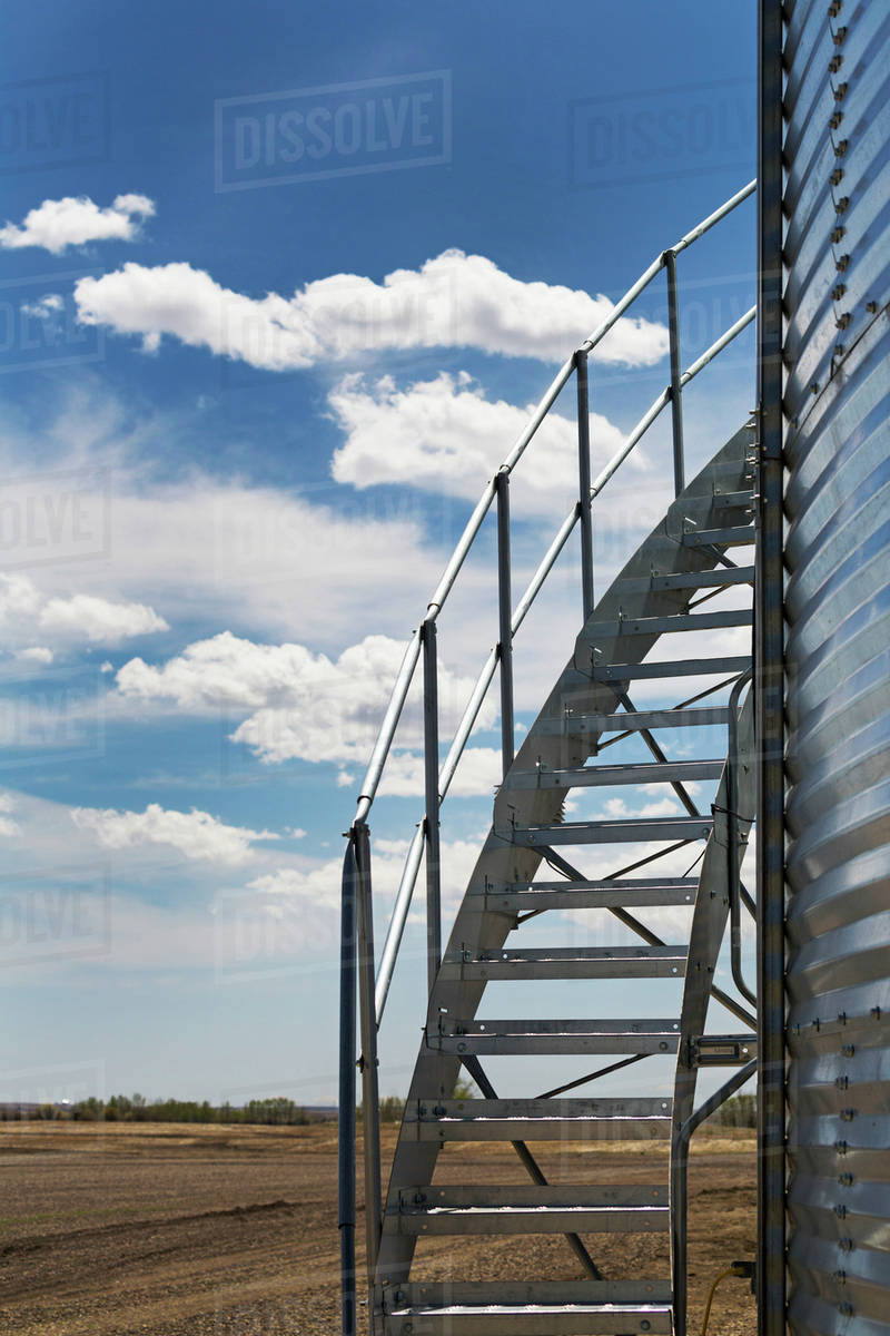 Close up of metal stair case on metal grain bin with clouds and blue ...