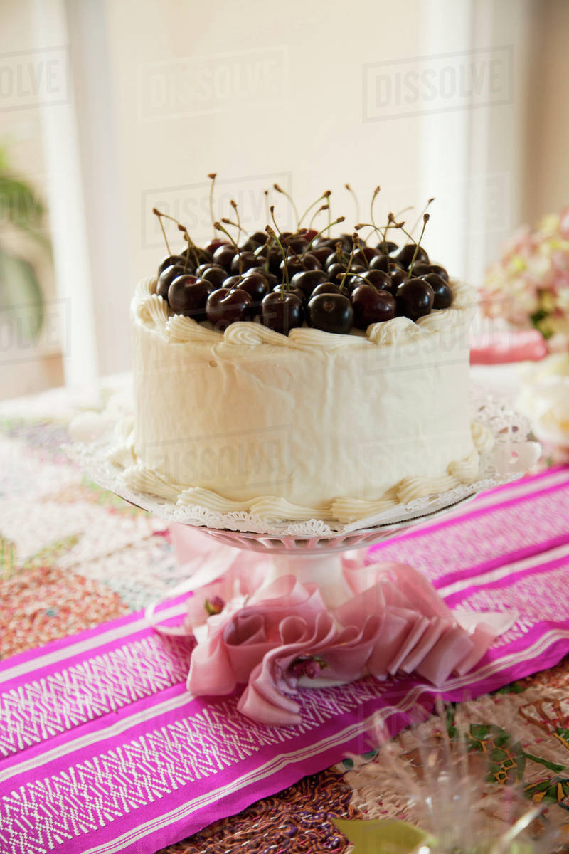 A white iced cake topped with fresh cherries on a pink table runner ...