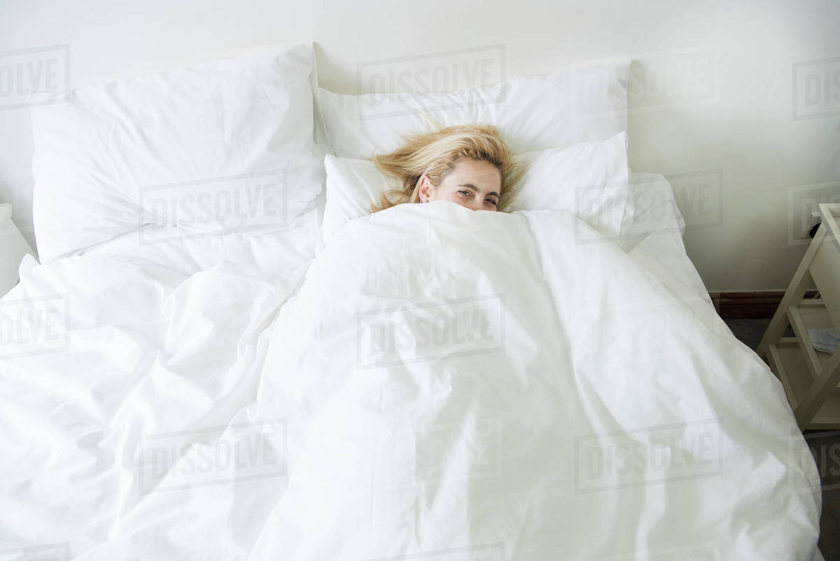 A cool young woman lying in a bed with he covers pulled up to her chin ...