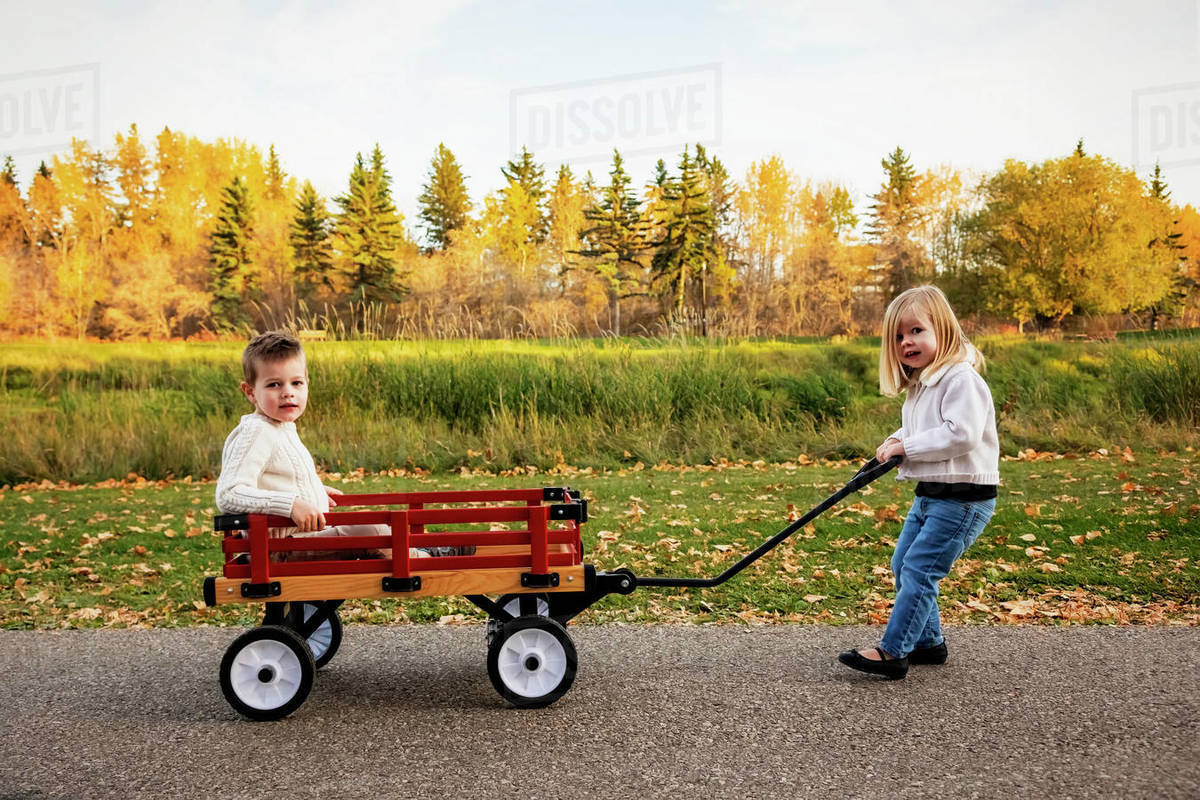 Young girl pulling her brother in a wagon at a city park along a river ...
