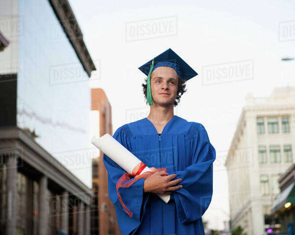 A graduate in cap and gown holding his degree on an urban street ...