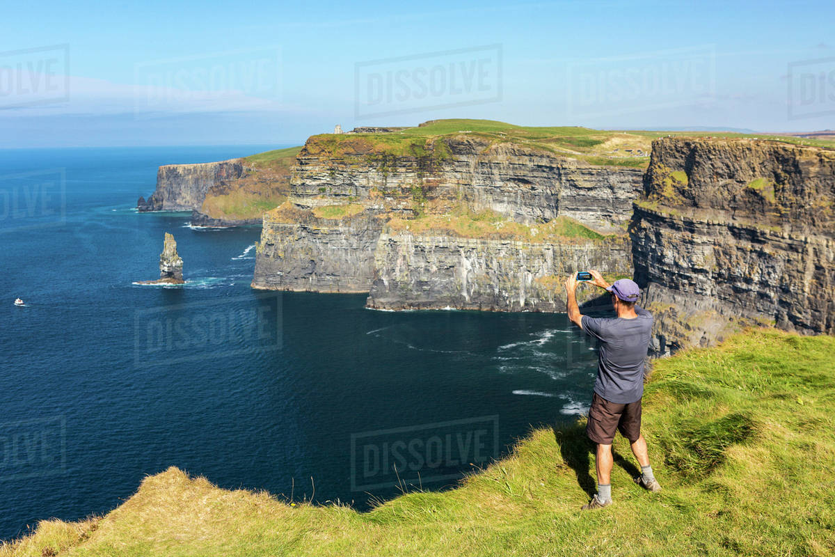 Man standing on grassy cliff's edge overlooking large sea stack rock ...