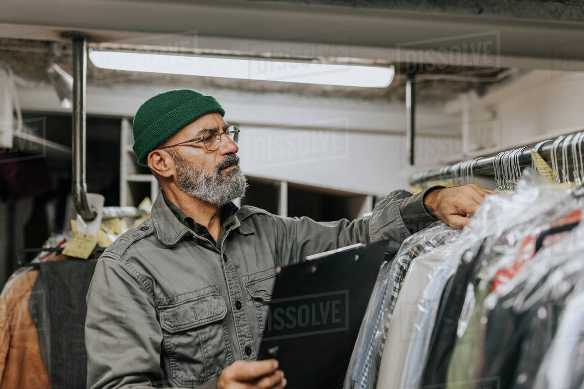Bearded male tailor with clipboard checking clothes hanging on rack in ...