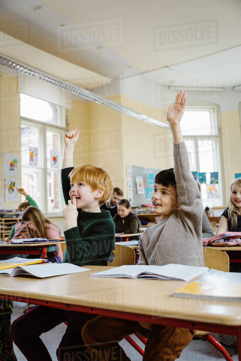 Smiling male pupils raising hands while answering in classroom - Stock ...
