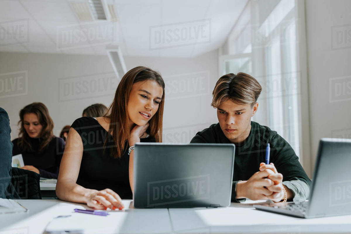 Multiracial male and female students sharing laptop at desk in ...