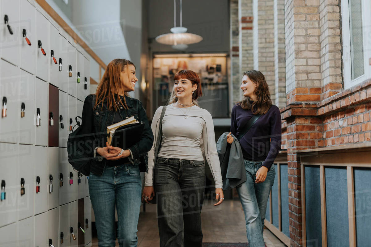 Happy female friends walking together by locker in corridor - Stock ...