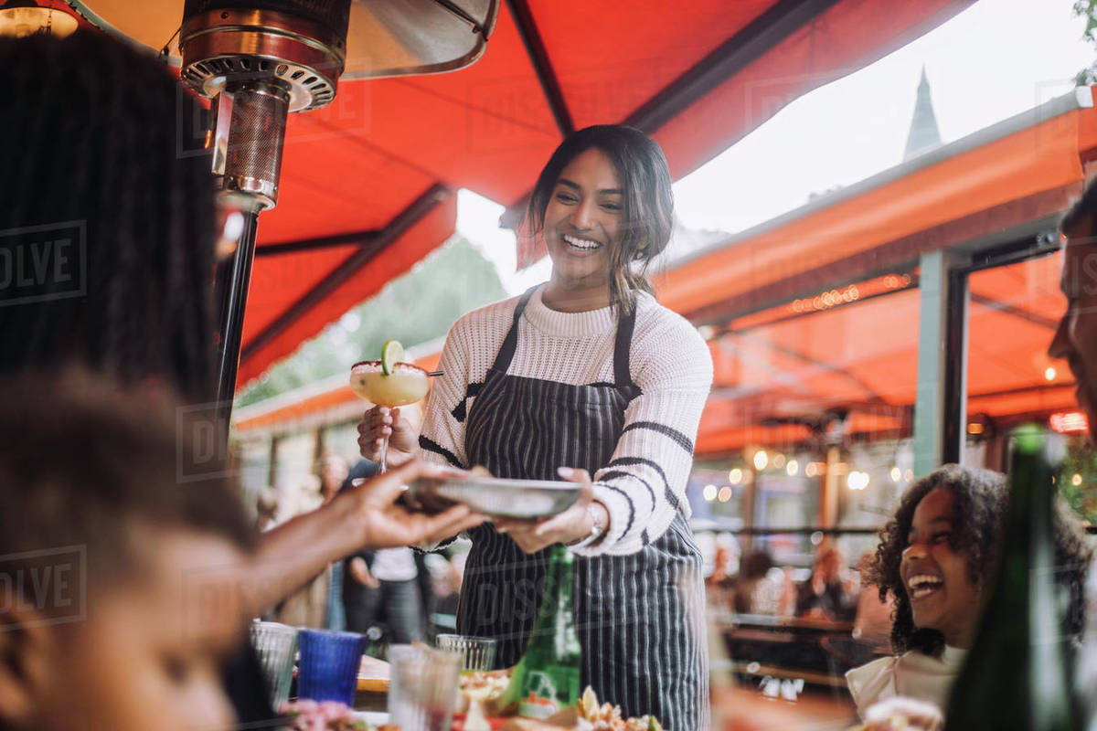 Smiling waitress serving food and drinks to customers having fun at ...