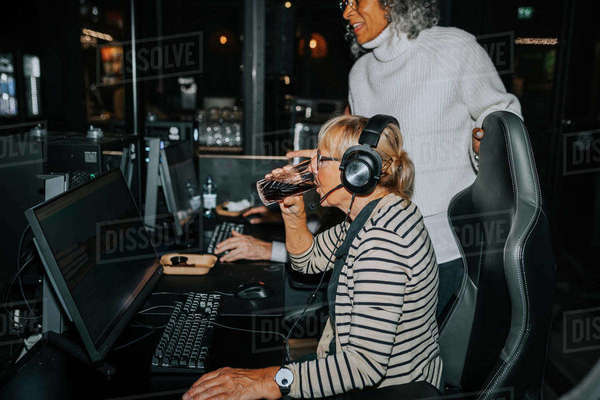 Elderly woman drinking water while playing computer game by female ...