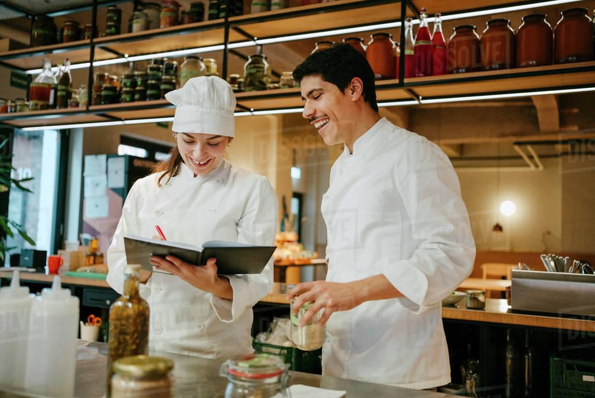 Happy female chef writing in diary with male coworker while discussing ...