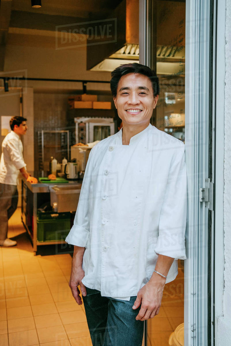 Portrait of happy male chef leaning on door at commercial kitchen ...