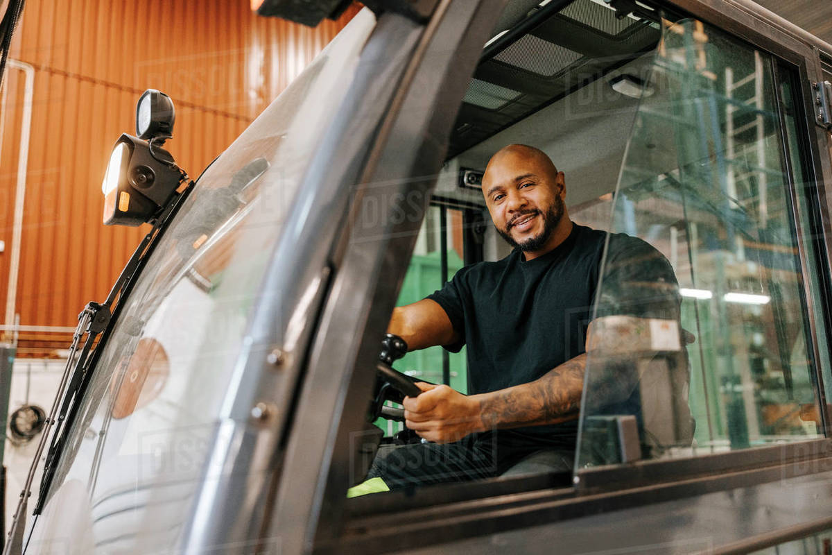 Portrait of smiling bald male worker sitting in forklift at factory ...