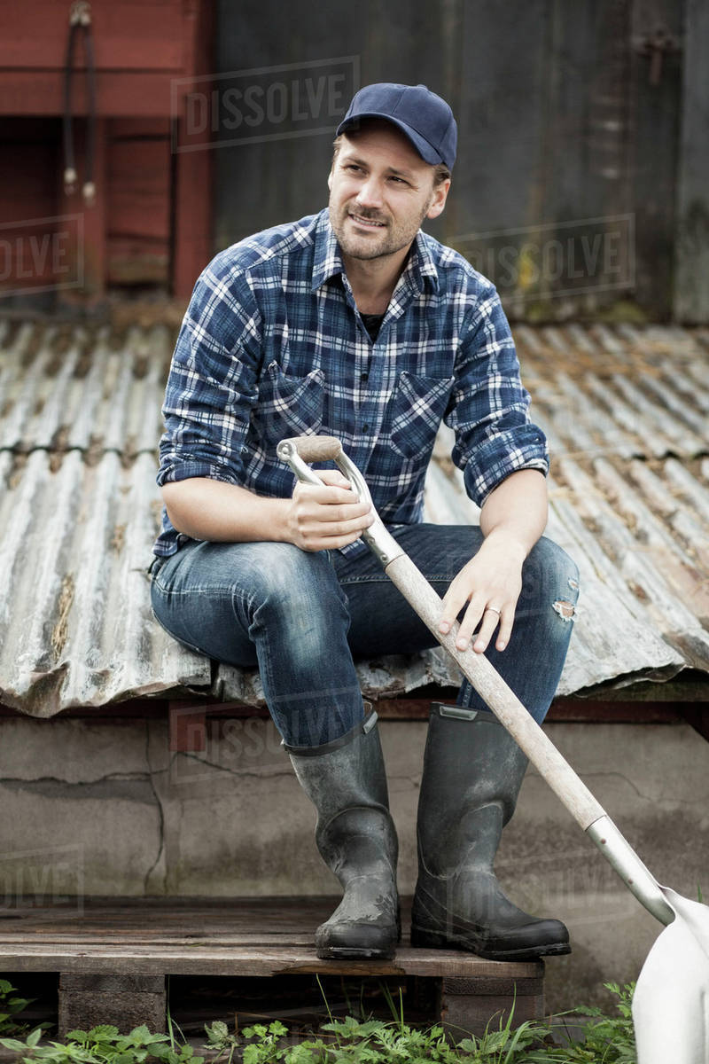 Full length of farmer with shovel sitting on corrugated iron at farm ...