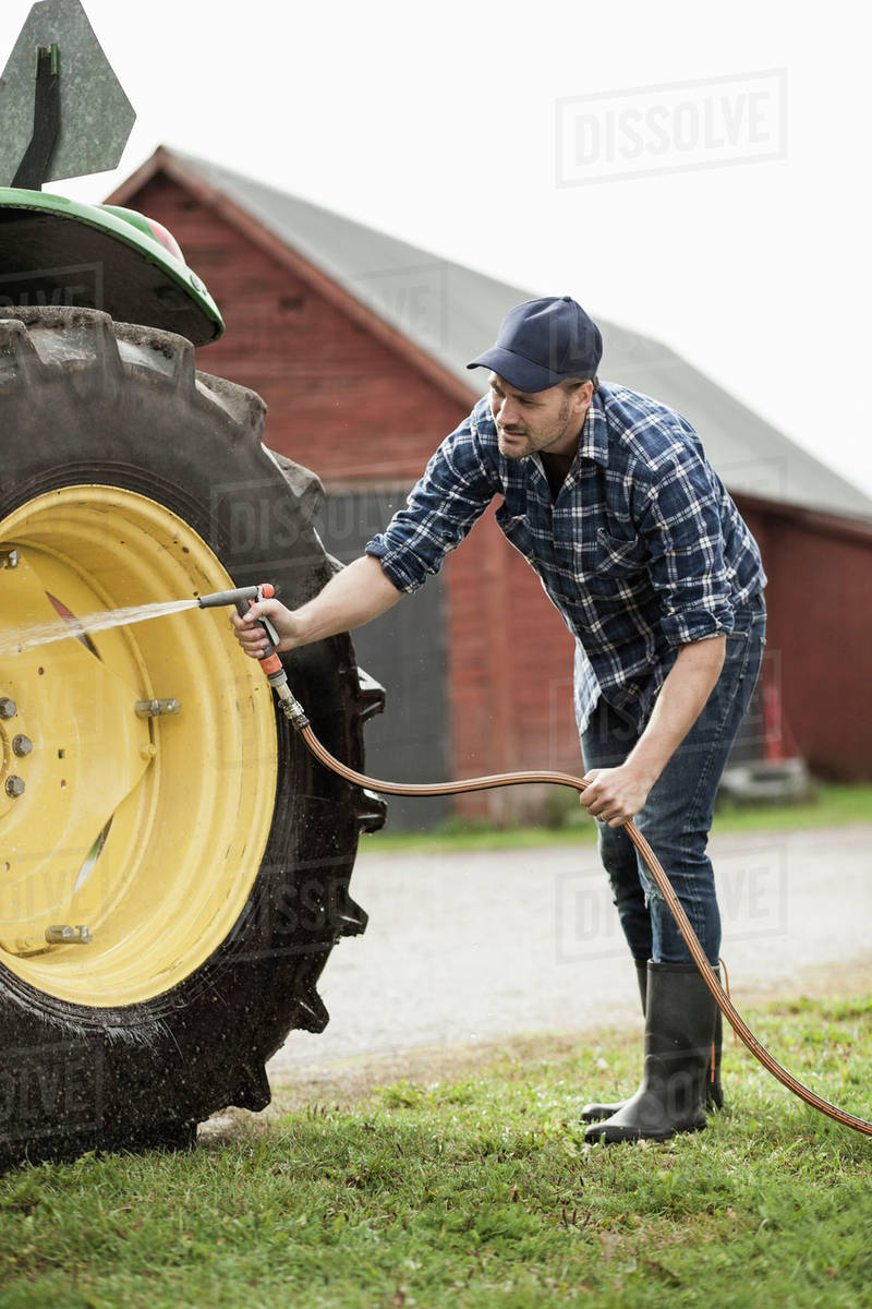 Full length mid adult man washing tractor wheel with hose in farm ...