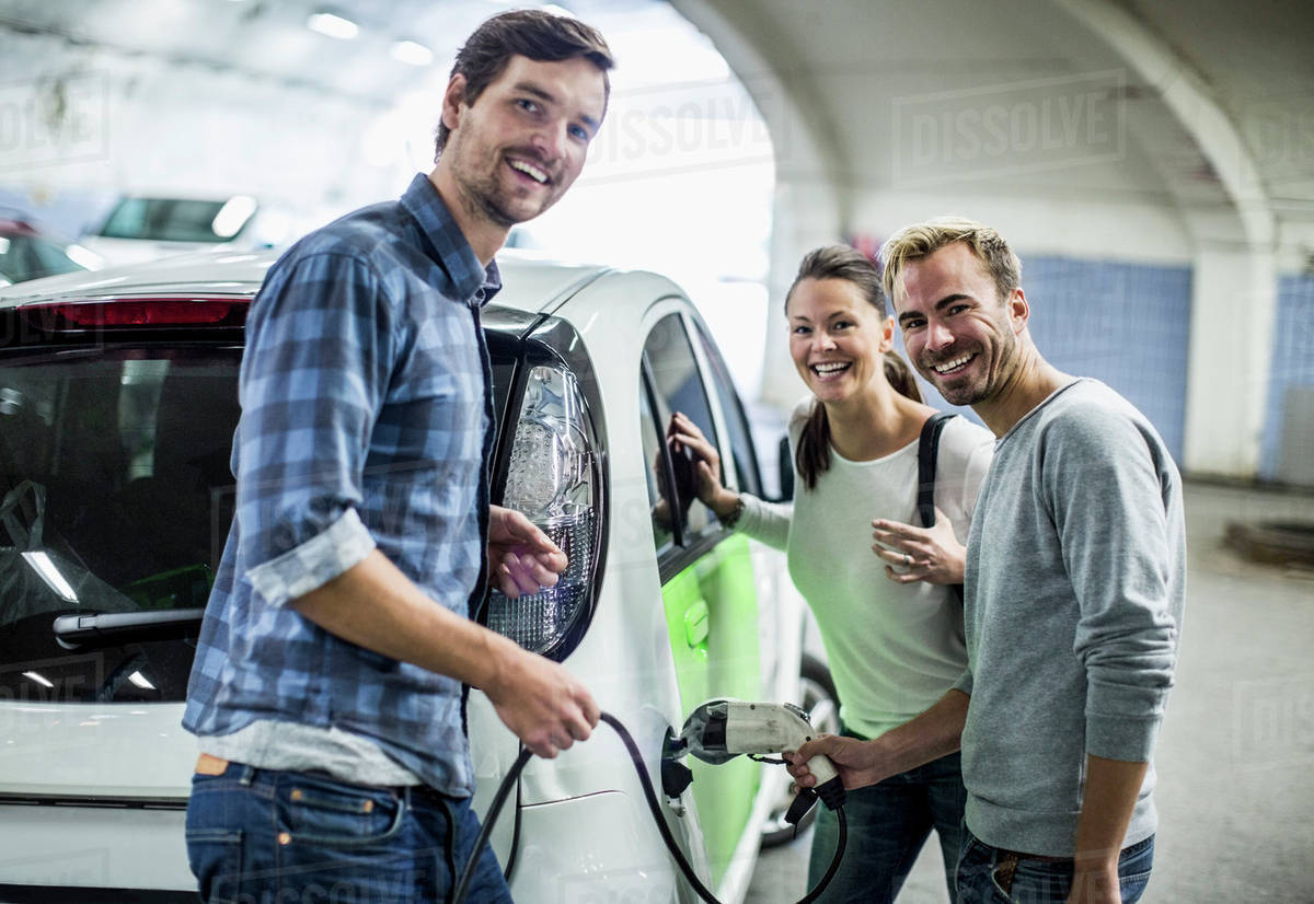 Portrait of happy friends with man charging electric car at gas station ...