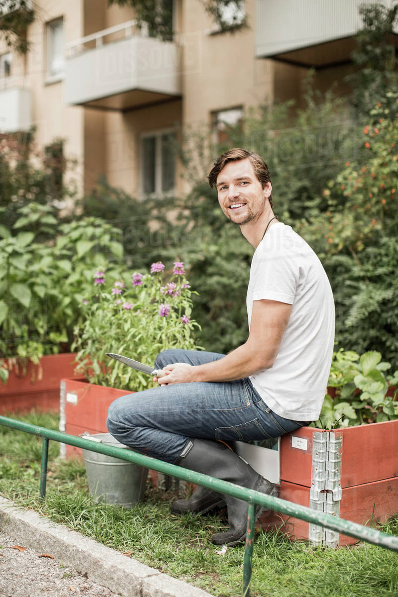 Full length portrait of young man with gardening equipment sitting in ...