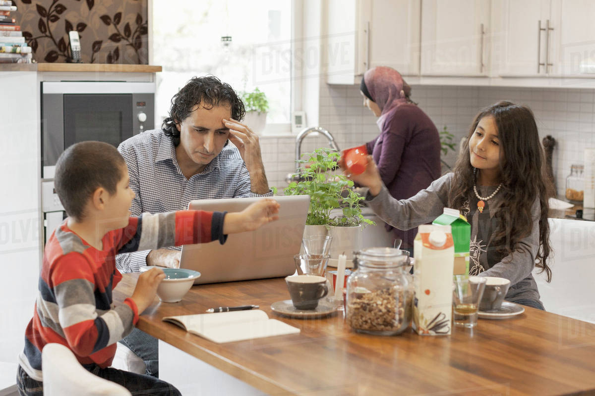 Muslim man working on laptop in kitchen with family having breakfast ...