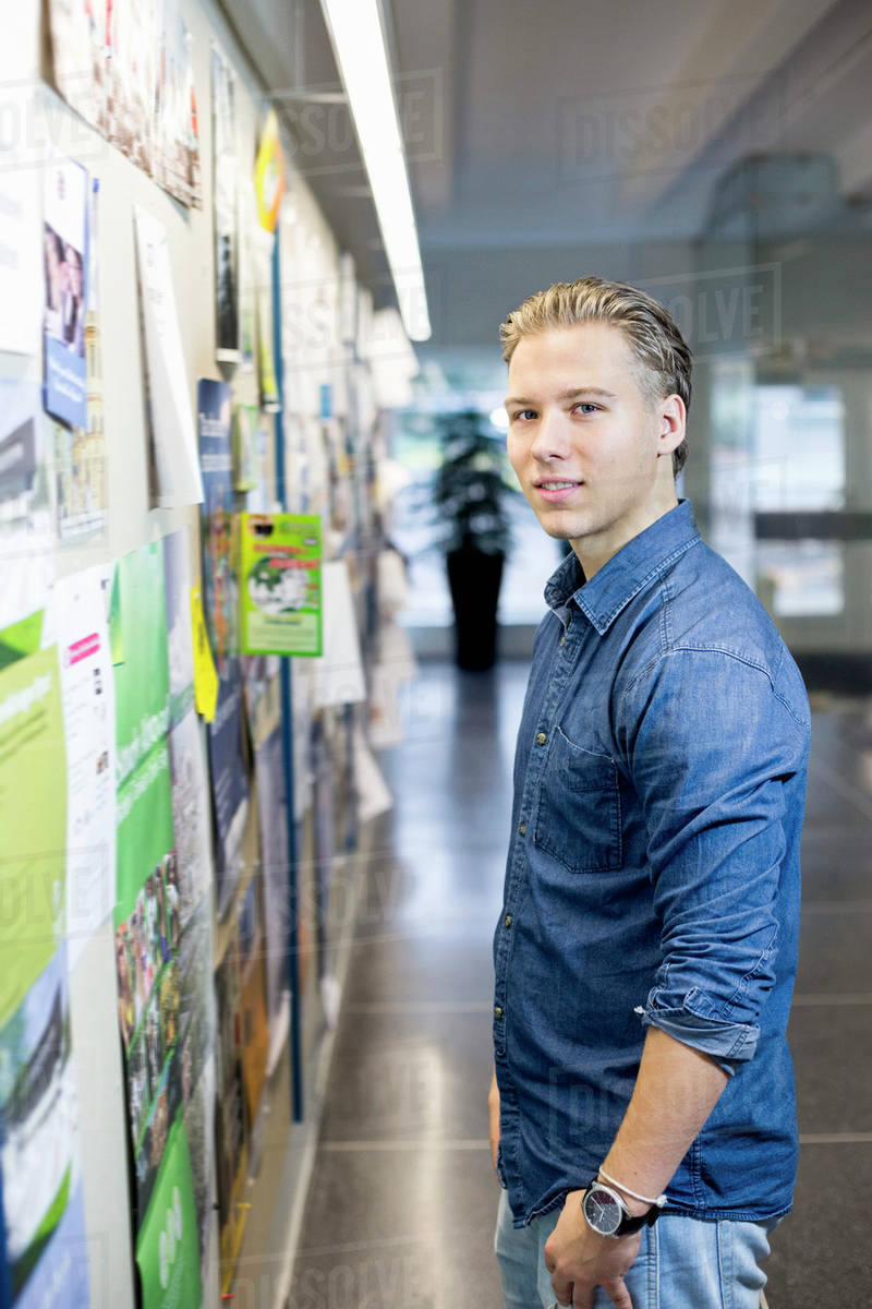 Portrait of young student standing in front of bulletin board at ...