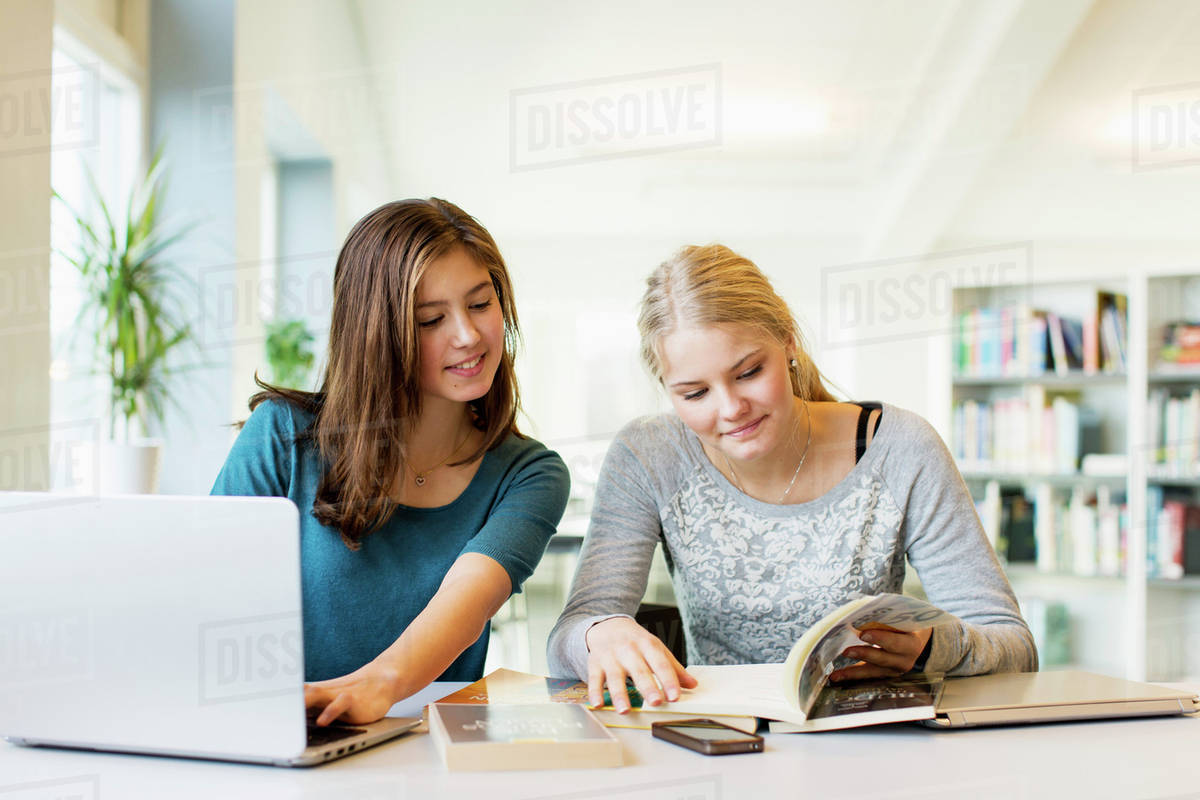 Teenage girls reading book while using laptop at table in school ...