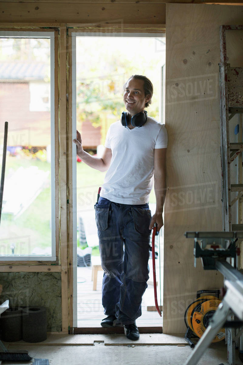 Full length of smiling man leaning in doorway of house being renovated ...