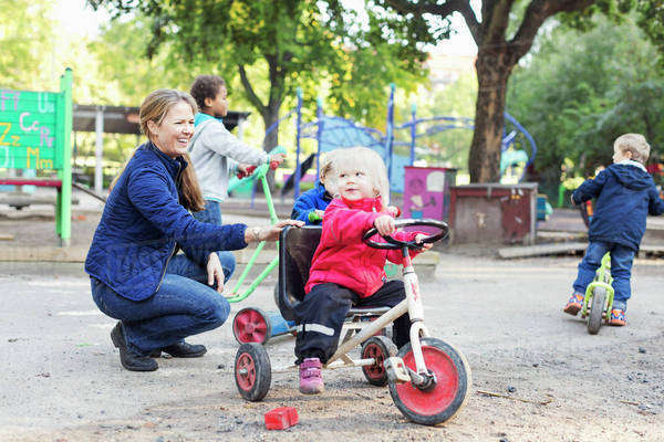 Happy female teacher assisting girl to ride tricycle on playground ...