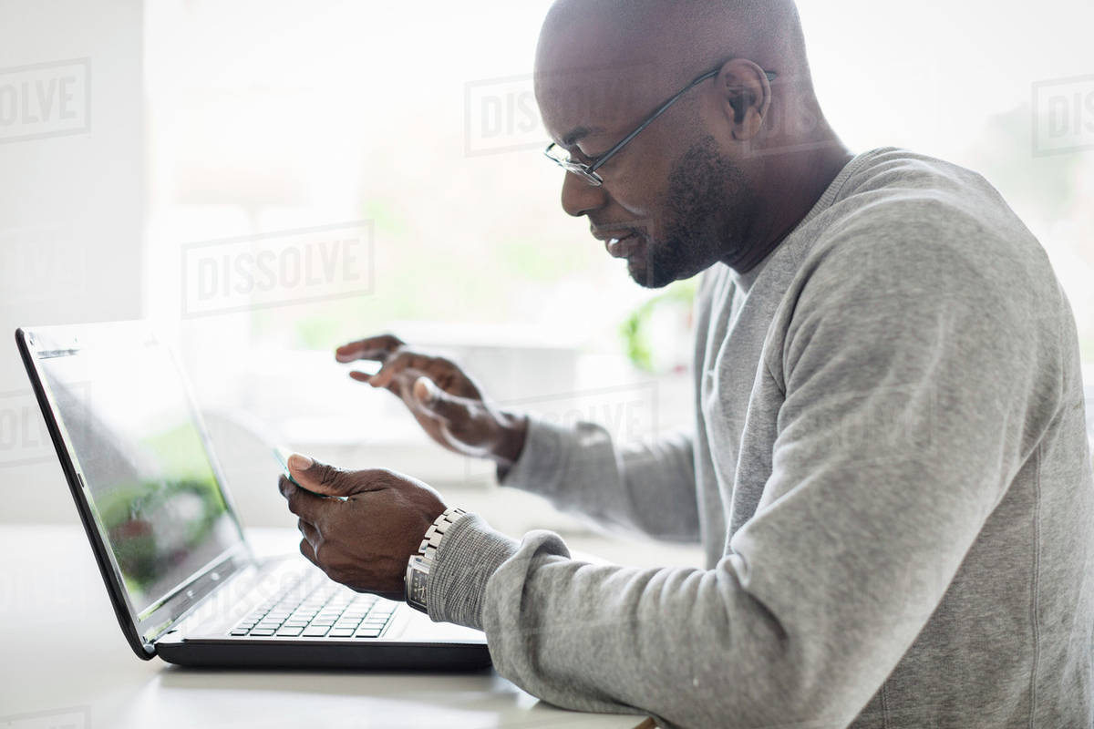 Side view of man using smart phone with laptop at table - Royalty-free ...