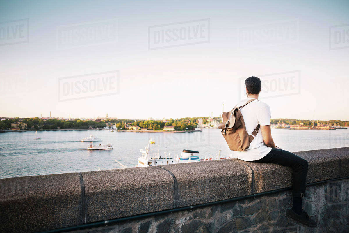 Male tourist looking at view while sitting on retaining wall of bridge ...