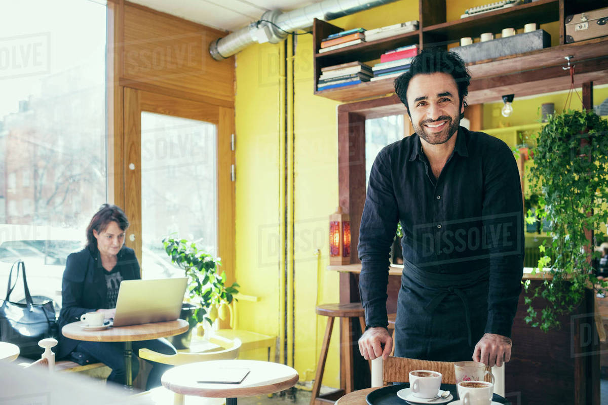 Portrait of happy waiter with customer using laptop in background at ...