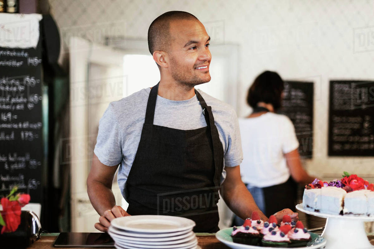 Smiling baker standing at cafe counter with coworker working in ...