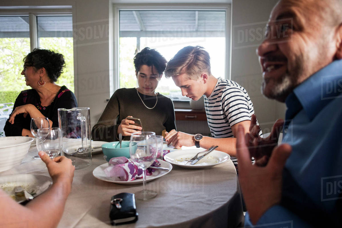 Friends using phone while family talking at dining table in dinner ...