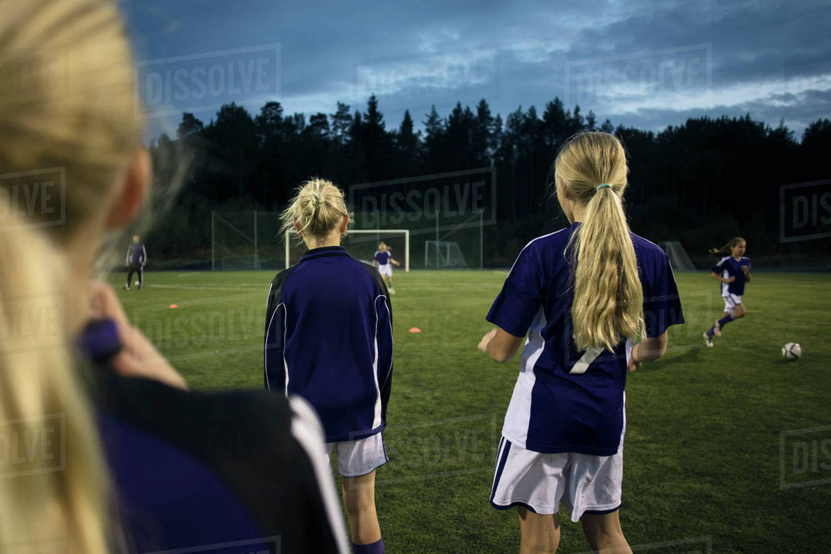 Rear view of girls standing on soccer field against sky at dusk ...