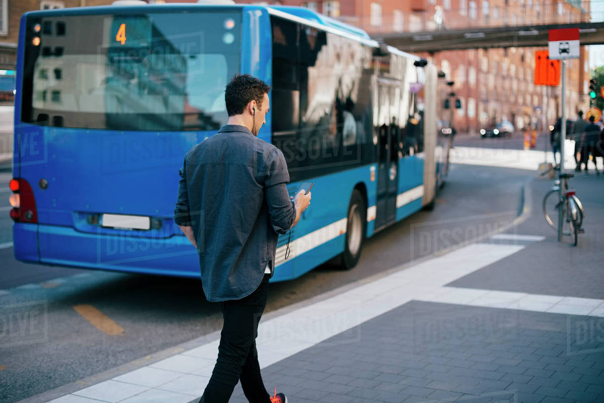 Rear view of mid adult man walking on sidewalk while using mobile phone ...