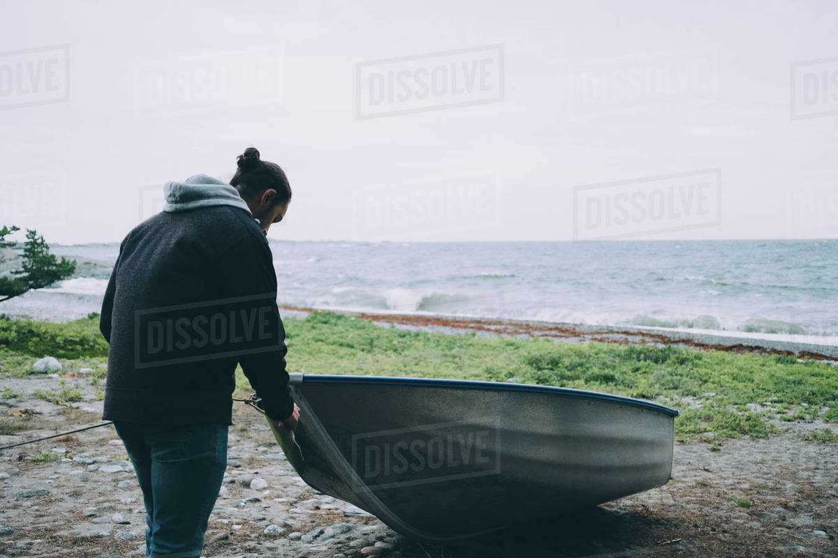 Rear view of young man standing by boat at beach - Stock Photo - Dissolve