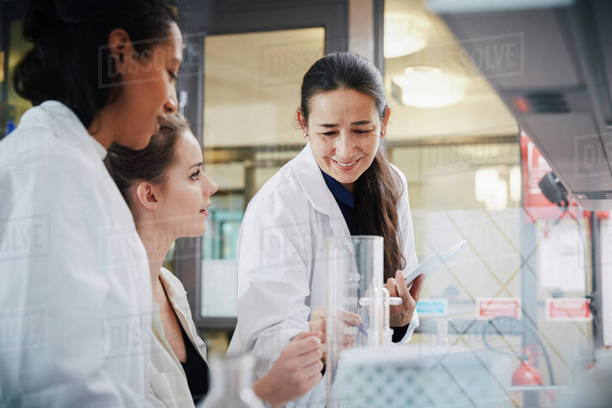 Smiling mature teacher with young female students learning chemistry at ...