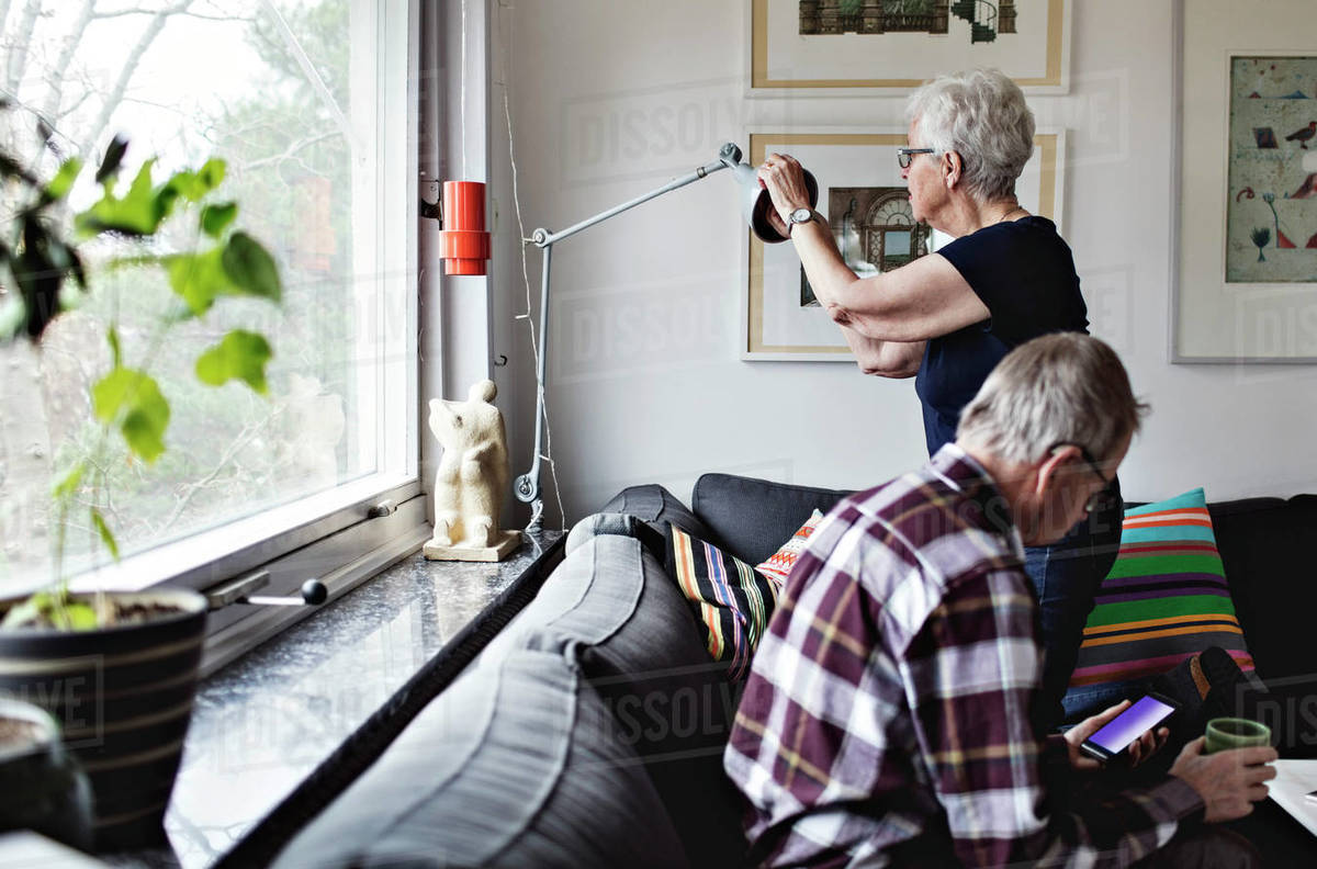 Senior woman adjusting floor lamp by man sitting on sofa in living room ...