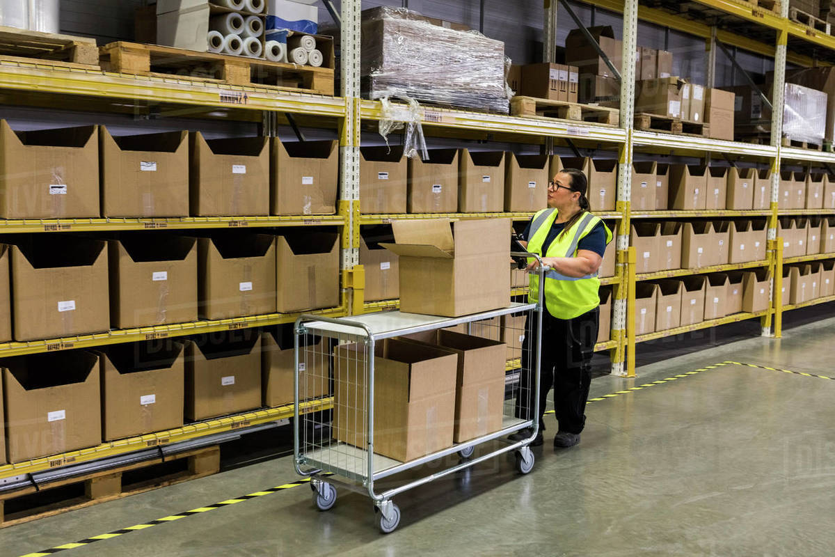 Full length of female worker pushing cart with cardboard boxes by rack ...