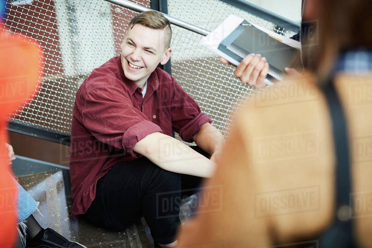 Smiling man sitting on steps while talking to friends in university ...
