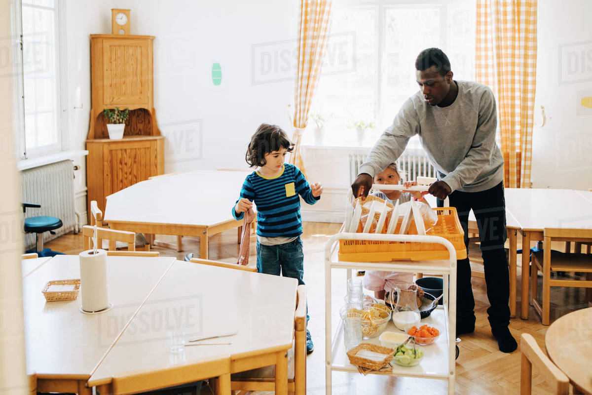 Male teacher assisting students pushing cart amidst tables in classroom ...