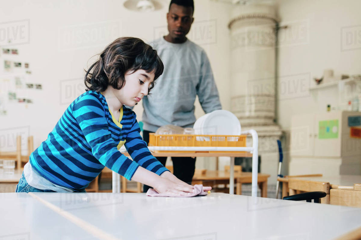 Boy cleaning table with dish cloth while teacher standing in classroom ...
