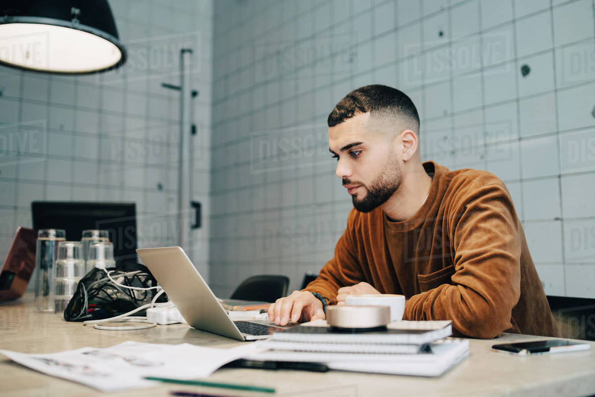 Confident young male computer programmer using laptop at desk in small ...