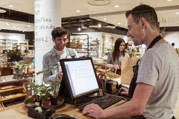 Cashier using computer while talking to smiling customer at checkout ...