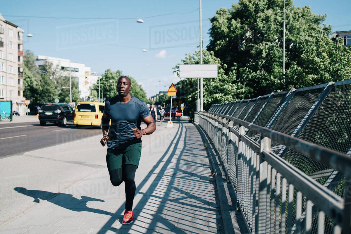 Full length of male athlete jogging on sidewalk at bridge in city ...