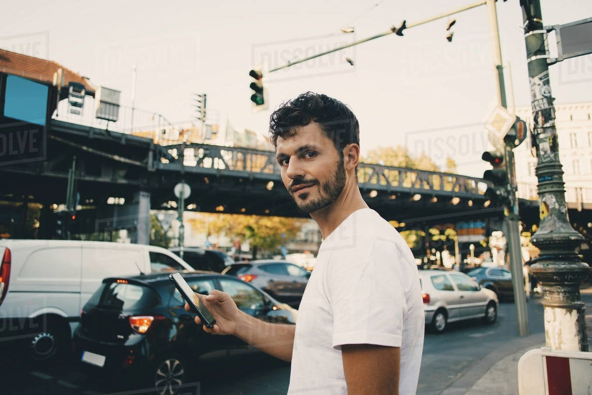 Portrait of young man holding mobile phone while standing on sidewalk ...