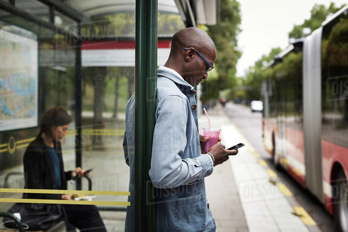 Businessman using smart phone while holding drink at bus stop in city ...