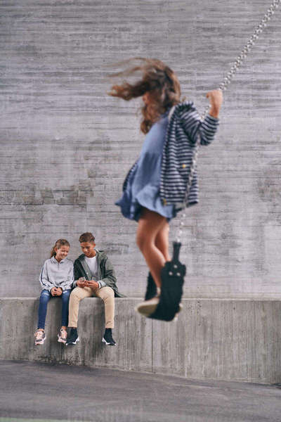 Full length of siblings using mobile phone while girl swinging on swing ...